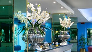 Uma área de recepção com balcão de mármore, dois vasos grandes com orquídeas brancas e um espaço de estar azul.