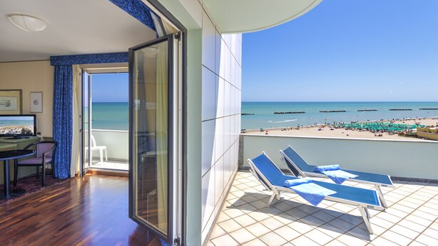 Uma varanda com vista para a praia, uma área de refeições com mesa e cadeiras, e uma TV em um suporte.