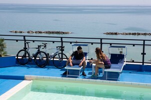Uma área à beira da piscina com duas espreguiçadeiras, uma bicicleta e vista para o mar.