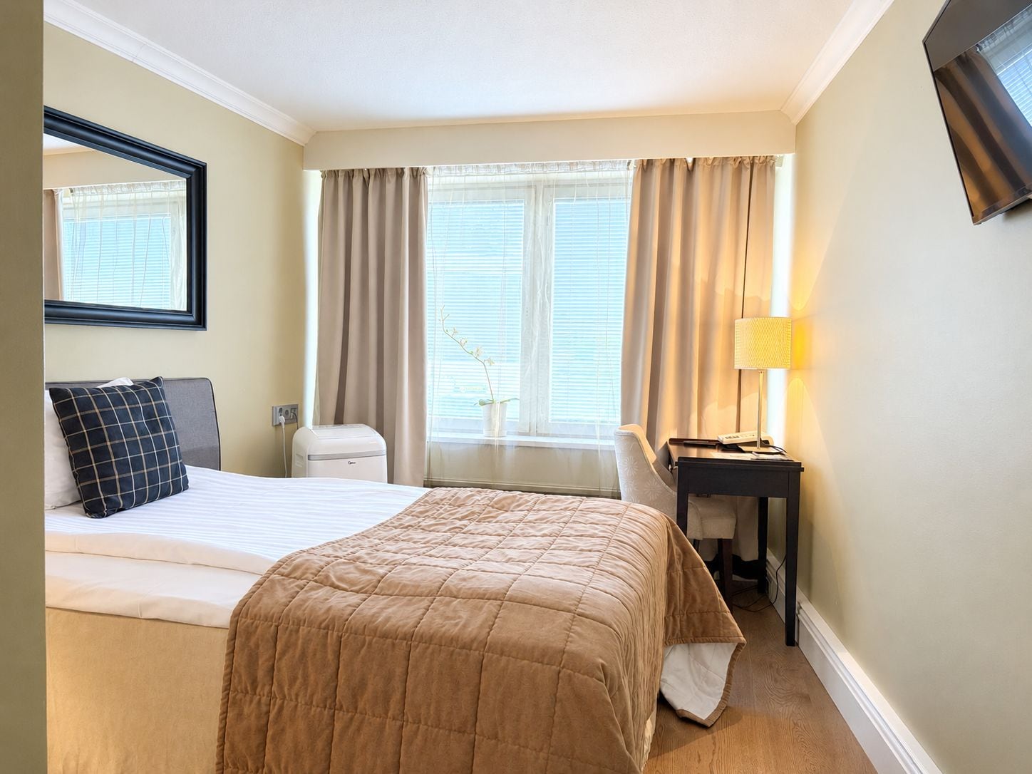 Quarto de hotel com cama, mesa de cabeceira, luminária, janela com cortinas e televisão presa na parede.