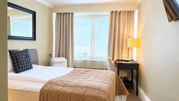 Quarto de hotel com cama, mesa de cabeceira, luminária, janela com cortinas e televisão presa na parede.