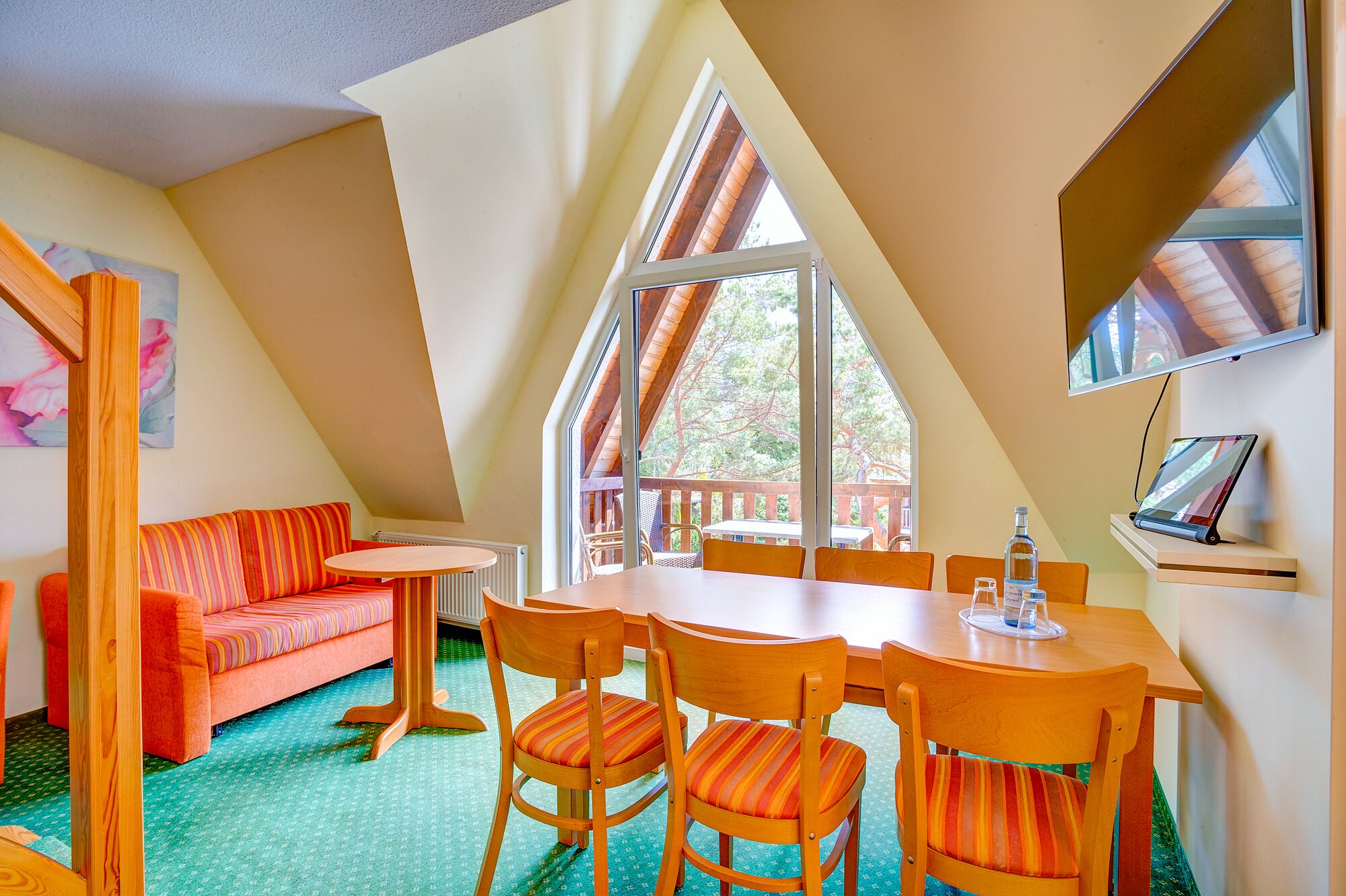 Um quarto aconchegante com teto inclinado, uma mesa de madeira com cadeiras, um sofá e uma janela grande com vista para árvores.