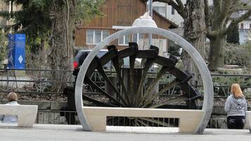 Uma grande escultura de metal com design circular, possivelmente uma roda d'água, em ambiente externo com bancos e árvores.