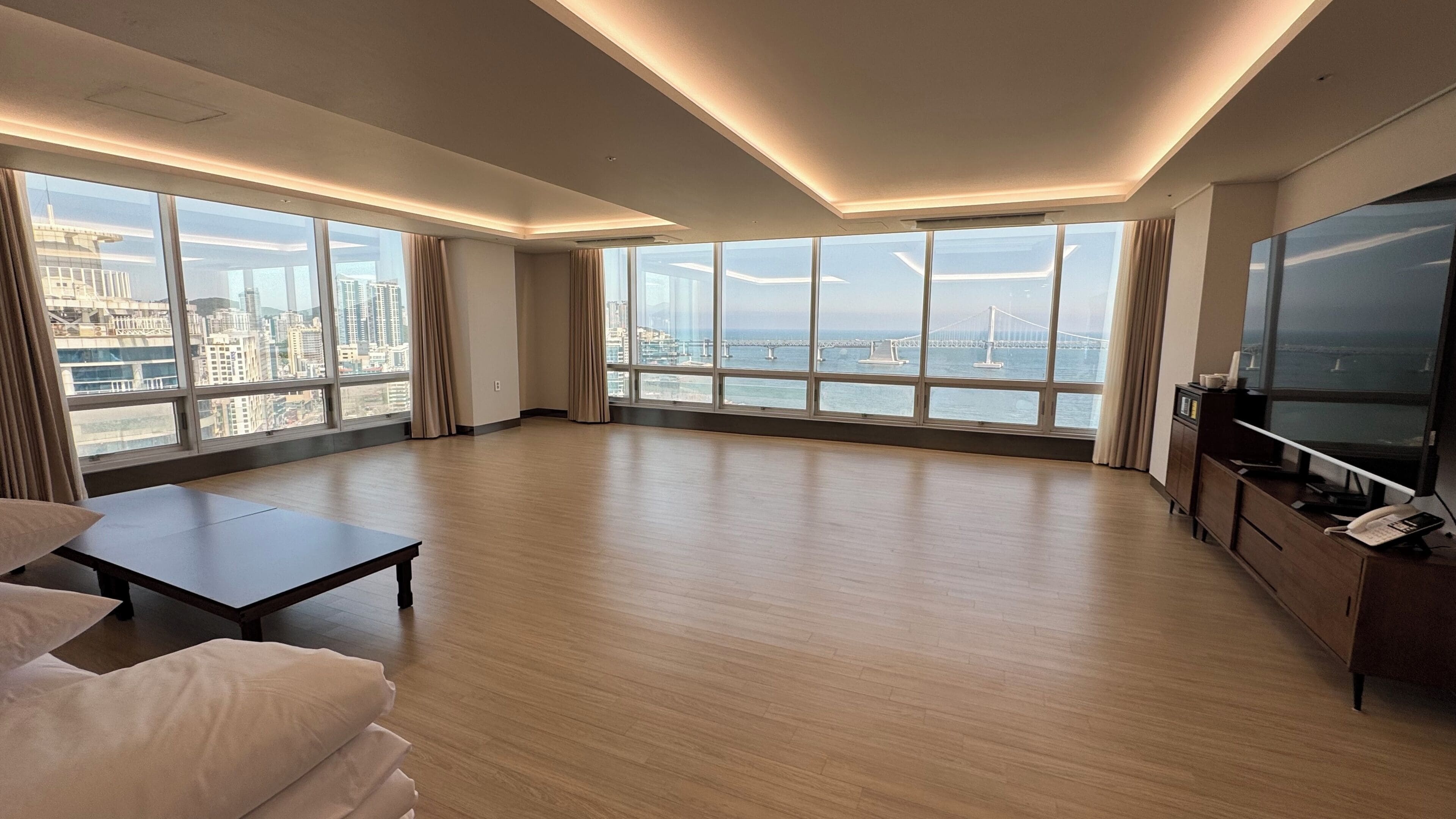 Ampla sala de estar com uma televisão grande, piso de madeira e vista para uma ponte e para a cidade através de grandes janelas.