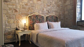 Quarto com parede de pedra, cama com cabeceira florida, mesa de cabeceira com abajur e uma janela.