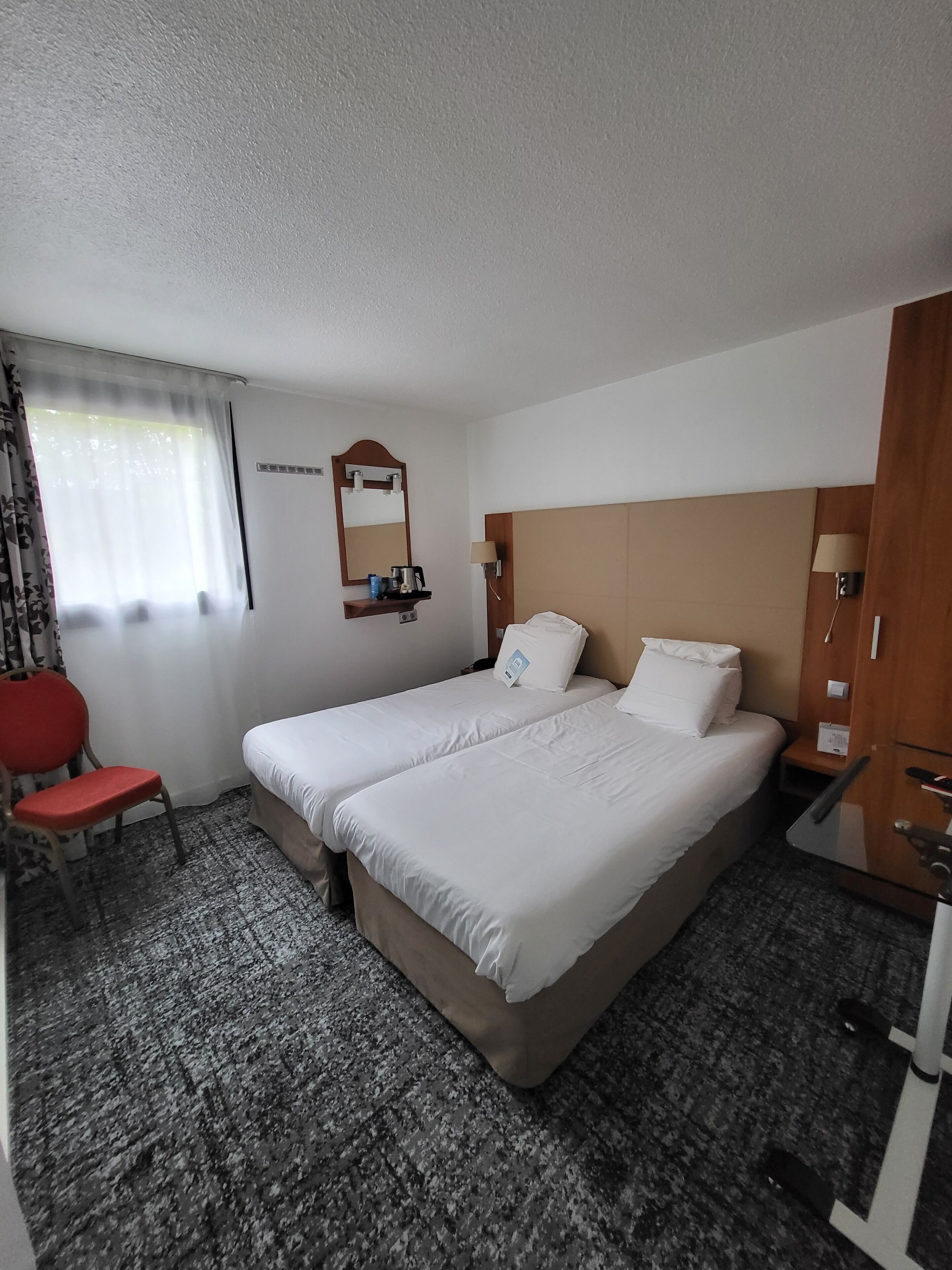 Quarto de hotel com duas camas, uma cadeira vermelha, um guarda-roupa de madeira, um espelho e uma janela com cortinas finas.