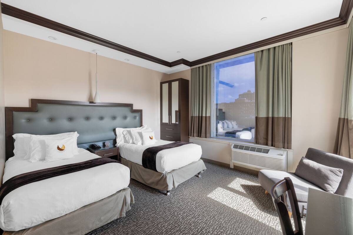 Quarto de hotel com duas camas, uma janela grande com cortinas, uma mesa e uma cadeira.