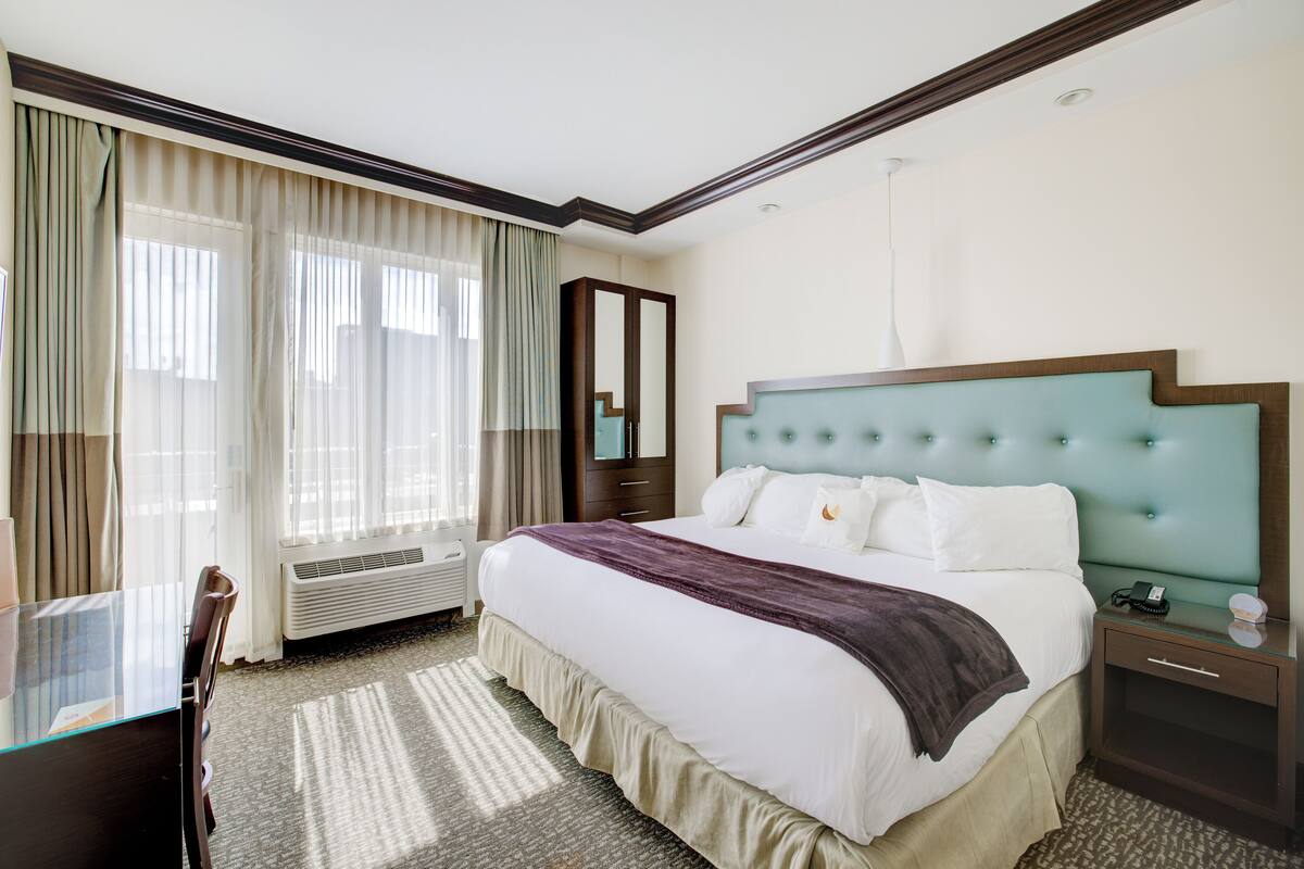 Quarto de hotel com uma cama grande, uma escrivaninha e uma cadeira. Há um espelho, uma mesa de cabeceira e um abajur.