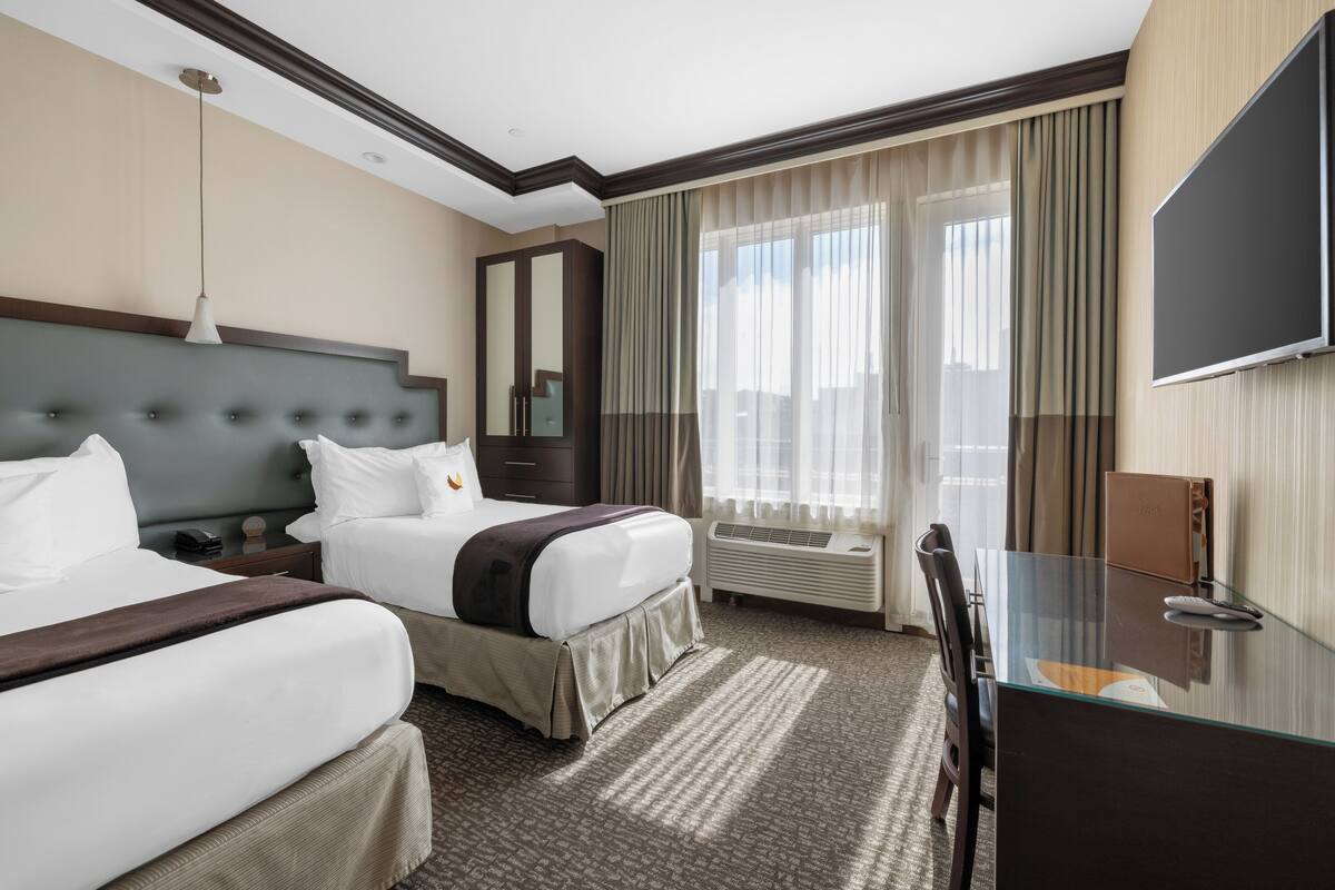 Quarto de hotel com duas camas, uma mesa, uma televisão e uma janela grande com cortinas.