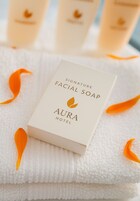 Uma caixa de Sabonete Facial Assinatura Aura Hotel, com caixa branca e texto dourado, colocada sobre uma toalha branca com fitas laranja.
