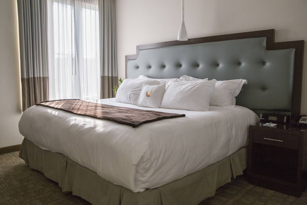 Uma cama bem arrumada com cabeceira estofada, mesa de cabeceira com abajur e janela com cortinas finas.