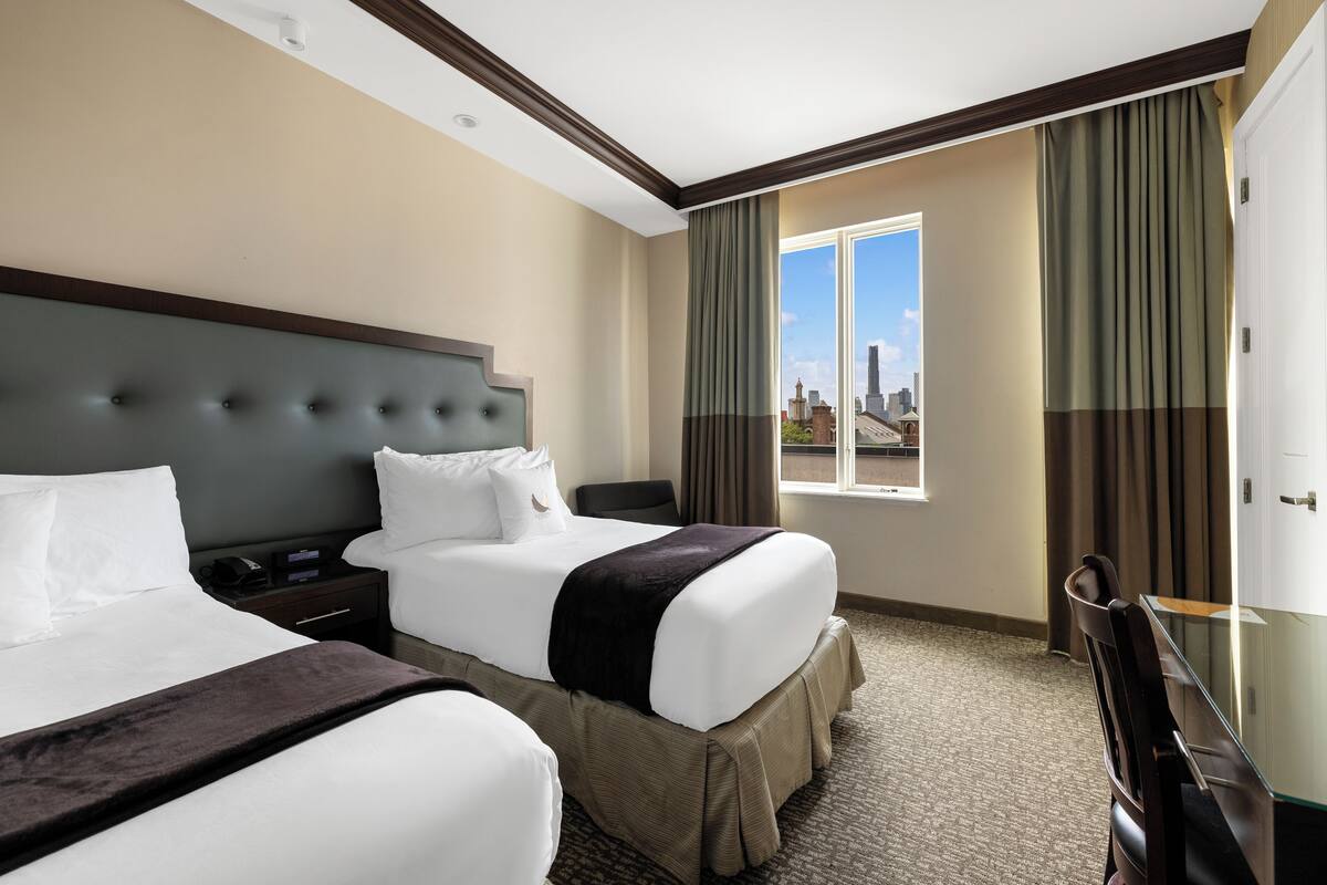 Quarto de hotel com duas camas, uma janela grande com vista para a cidade, uma escrivaninha e uma cadeira.