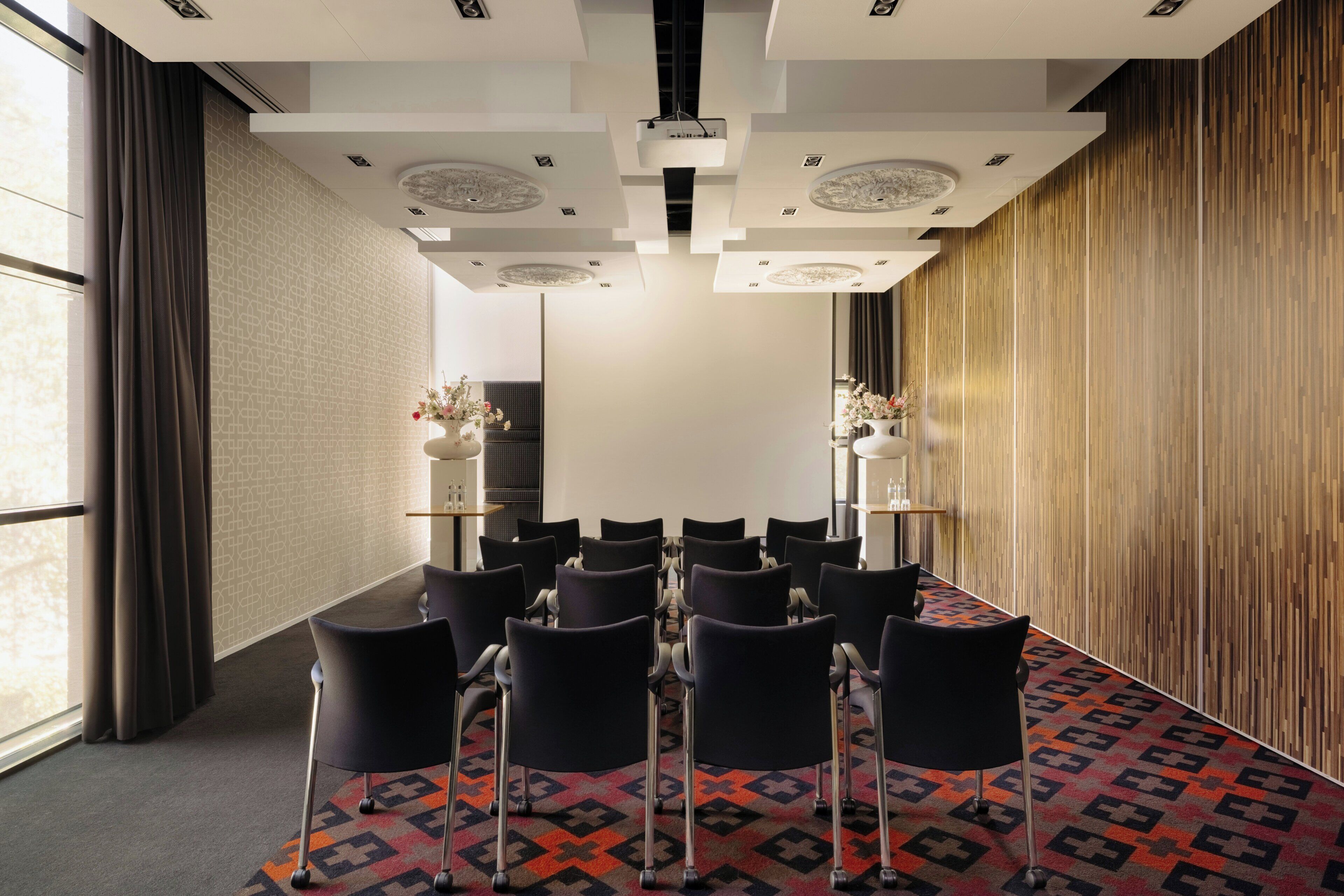 Uma sala de conferências com uma mesa comprida, cadeiras pretas, carpete estampado e parede de madeira.