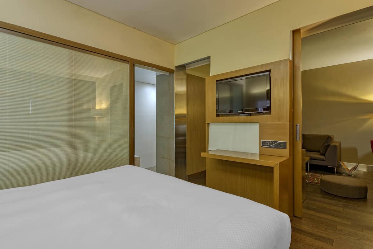 Quarto de hotel moderno com uma cama grande, uma TV de tela plana presa a um console de madeira, um espaço de estar com sofá e apoio para os pés, e uma porta de vidro deslizante que dá para um banheiro.