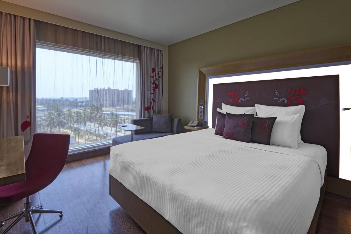 Quarto de hotel com uma cama grande, uma escrivaninha, uma cadeira e vista para palmeiras e edifícios.