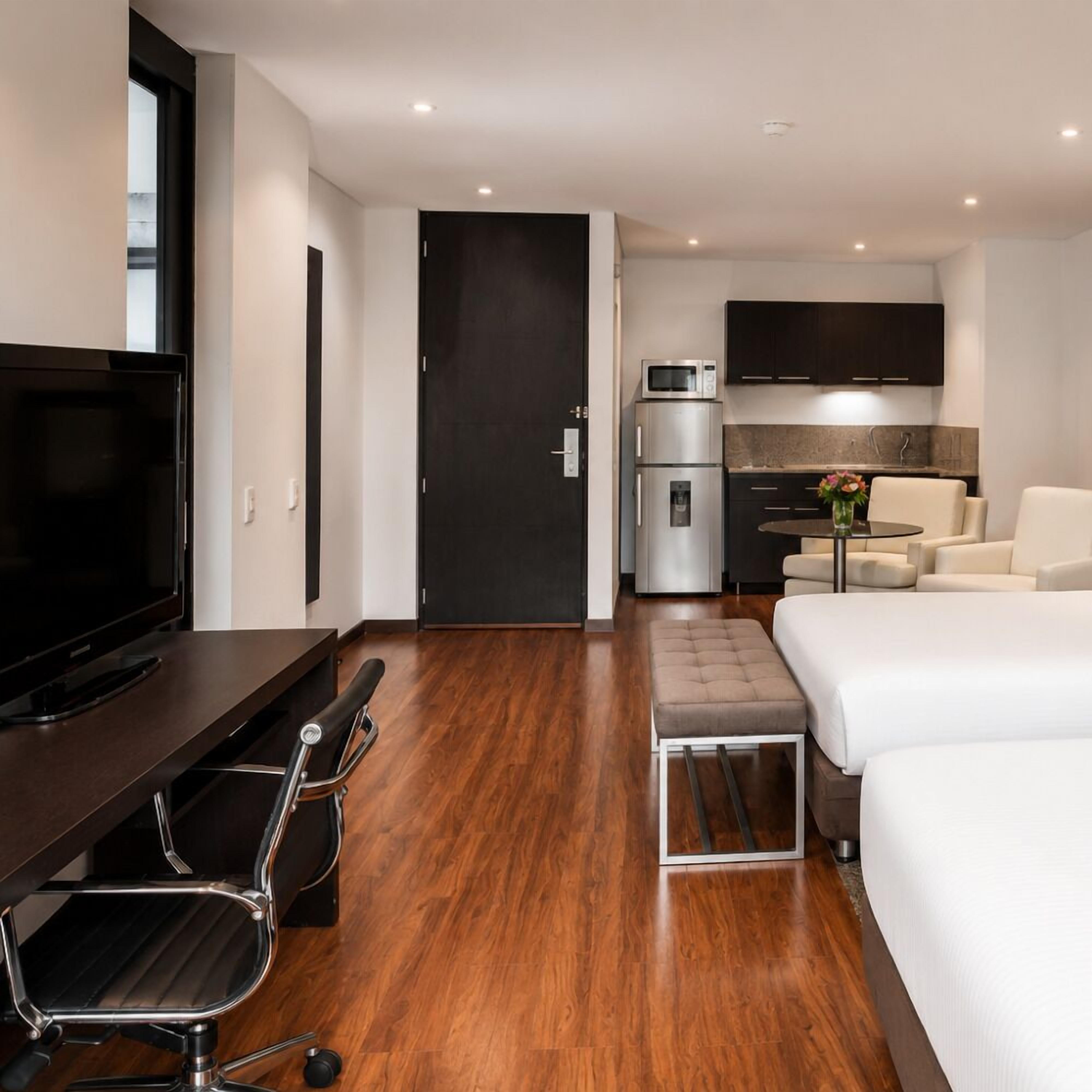 Quarto de hotel moderno com televisão de tela plana, escrivaninha com cadeira, sofá, mesinha com vaso e pequena cozinha no fundo.