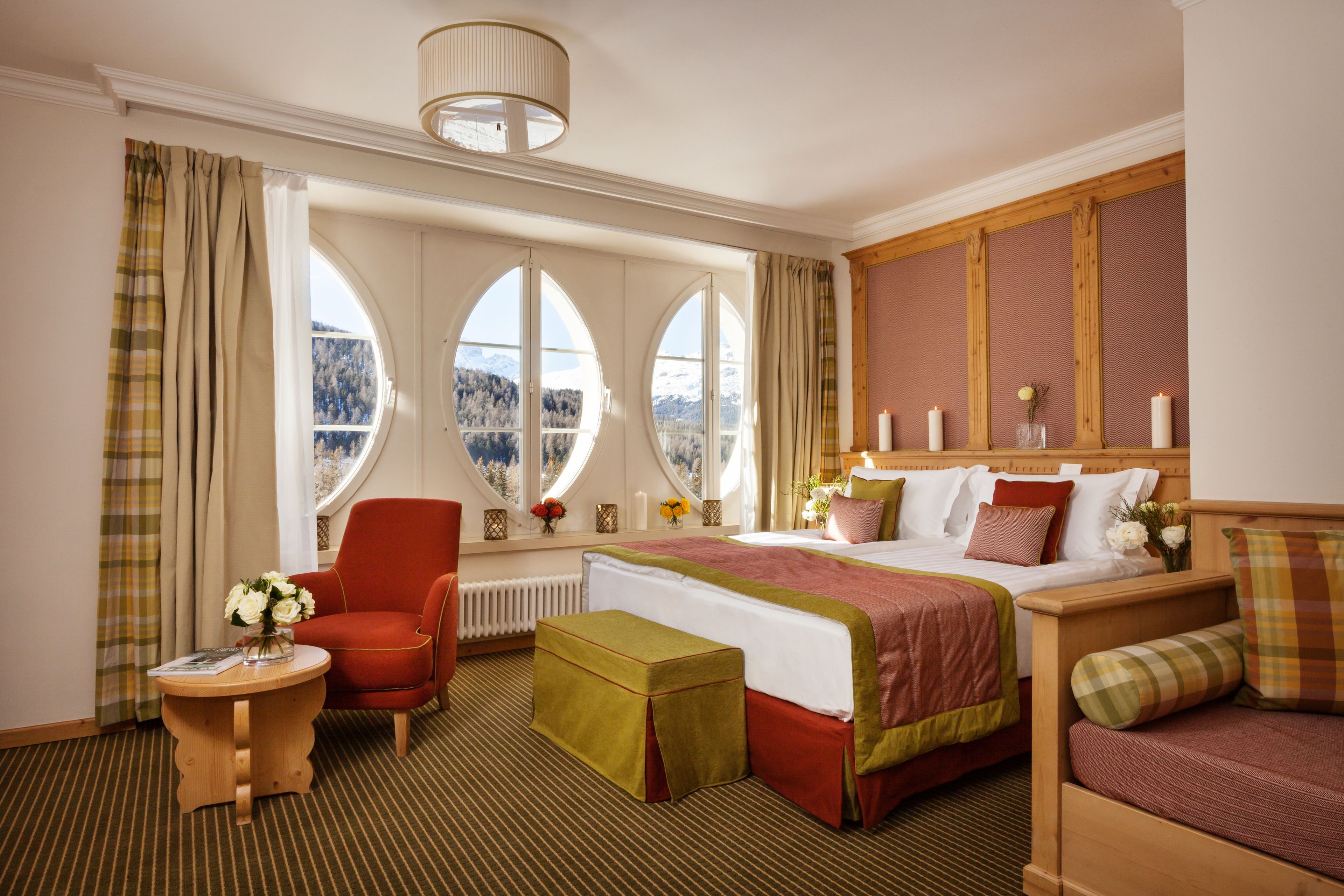 Quarto de hotel com uma cama grande, uma cadeira, uma mesinha com flores e vista para montanhas através de janelas amplas.