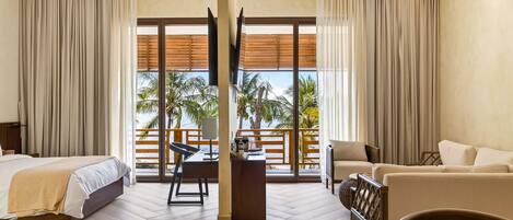 Quarto de hotel com cama, escrivaninha, cadeira, sofá e janelas amplas com vista para palmeiras e o mar.