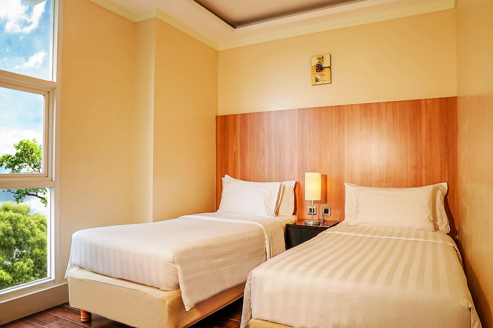Quarto de hotel com duas camas, cabeceira de madeira, mesa de cabeceira com abajur e janela com vista para árvores.