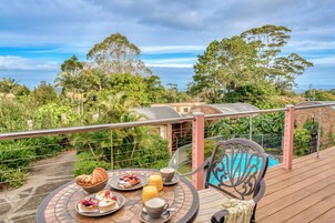 Uma mesa de jantar com itens de café da manhã, uma cadeira e vista para um jardim exuberante e edifícios ao longe.