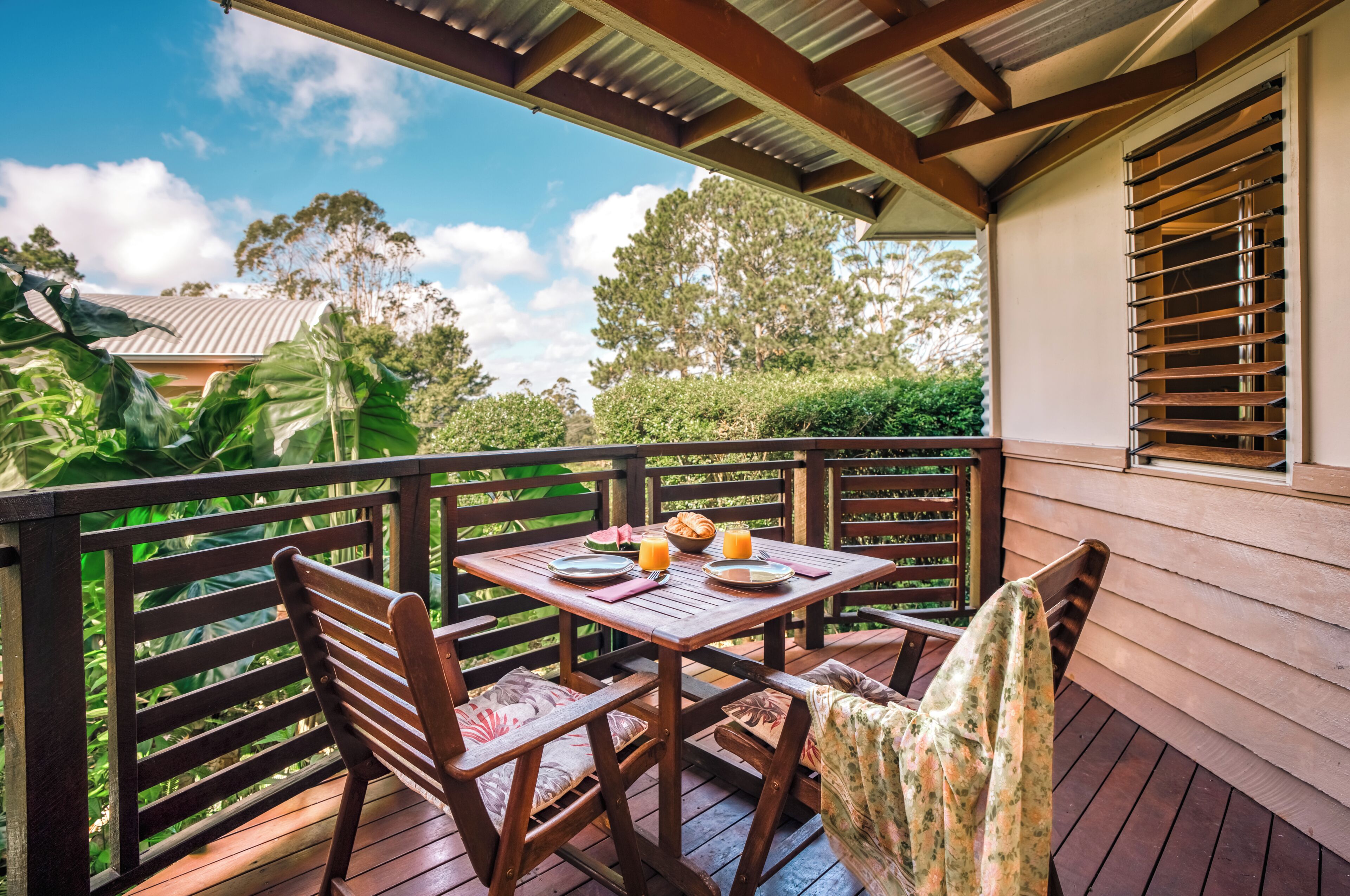 Um deck de madeira com uma mesa posta para dois, rodeado por cadeiras de madeira, com vista para um jardim.