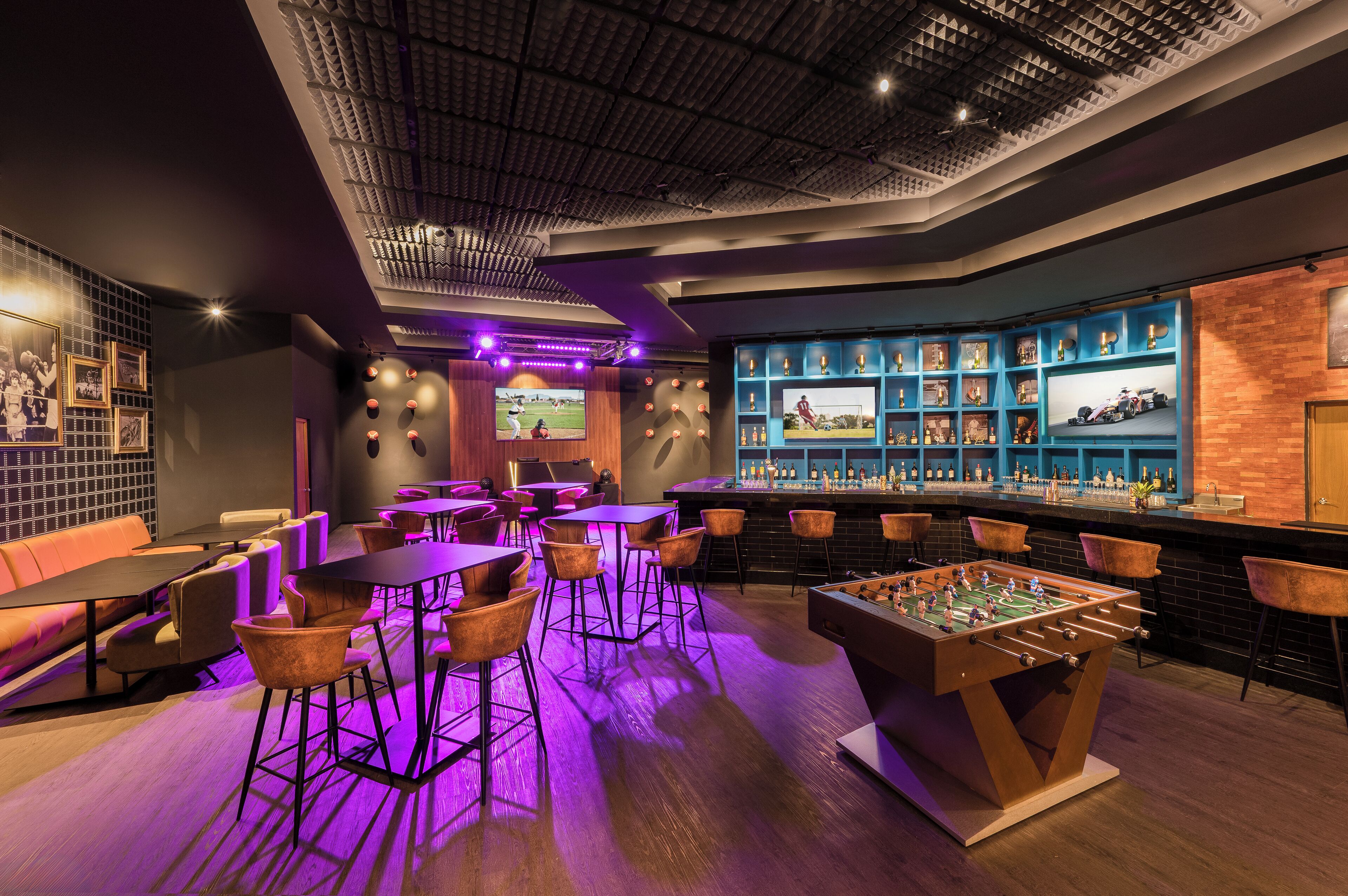 Uma área de bar com banquetas, uma mesa de pebolim e prateleiras com garrafas.