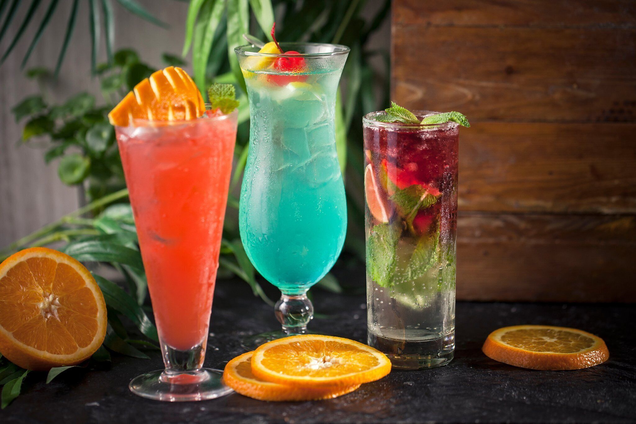 Três bebidas diferentes com guarnições de frutas e uma fatia de laranja sobre uma superfície escura.