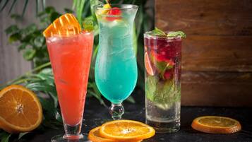 Três bebidas diferentes com guarnições de frutas e uma fatia de laranja sobre uma superfície escura.