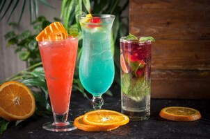 Três bebidas diferentes com guarnições de frutas e uma fatia de laranja sobre uma superfície escura.