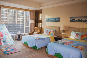 Quarto de hotel com duas camas, uma cadeira, um abajur, uma janela com vista para a paisagem urbana e uma tenda com personagens de desenho animado.