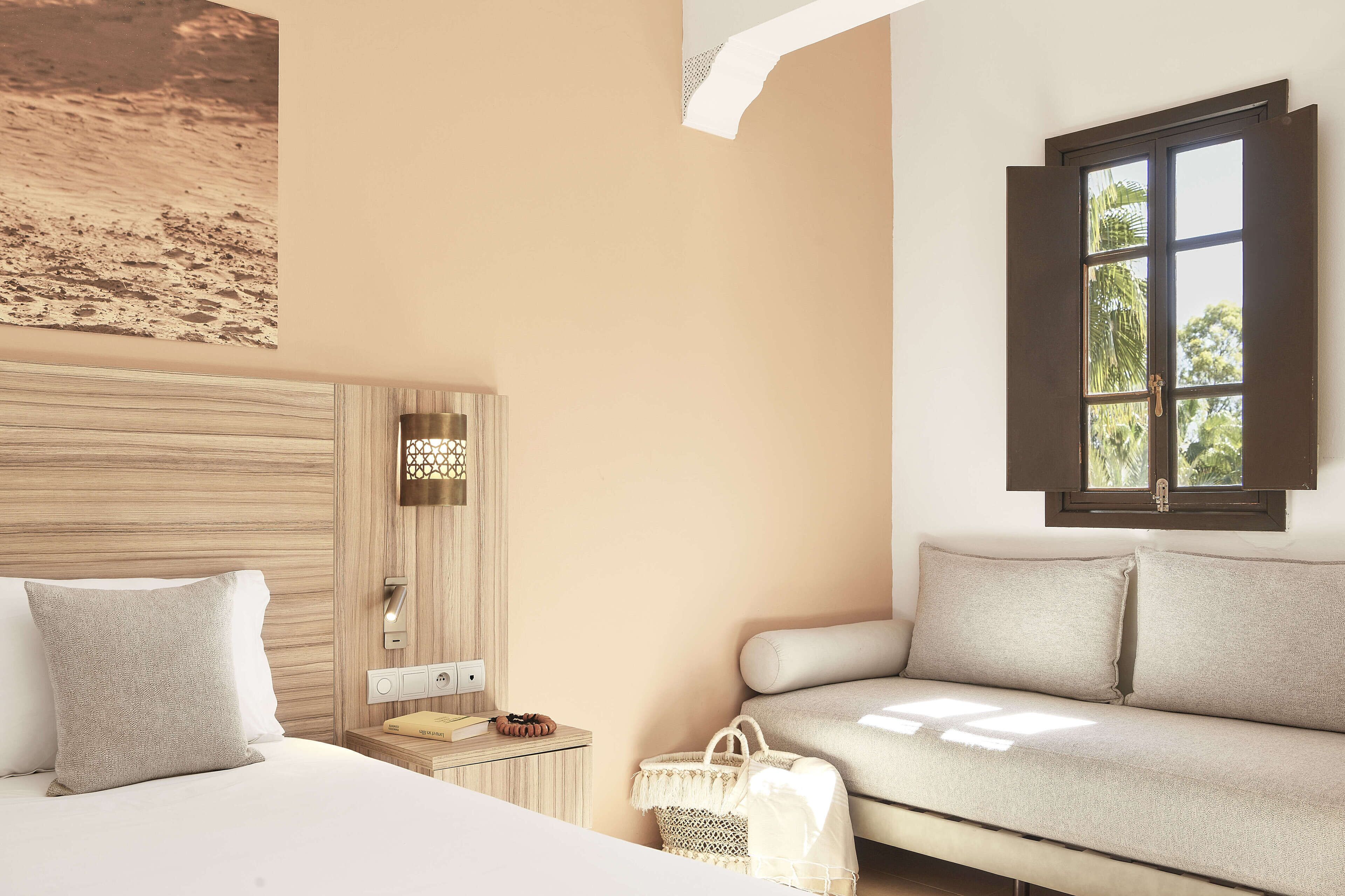 Quarto de hotel com cama, sofá, mesa de cabeceira, luminária e janela com vista para árvores.