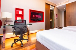Quarto de hotel com uma cama, uma escrivaninha com cadeira, uma televisão na parede e uma prateleira vermelha com um livro.