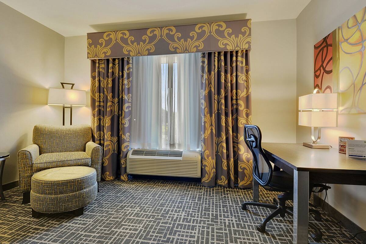Quarto de hotel com sofá, poltrona, apoio para os pés, escrivaninha e ar condicionado.