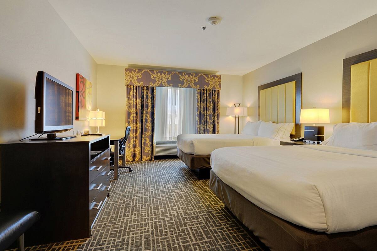 Quarto de hotel com duas camas, uma mesa com computador, televisão e janela com cortinas.