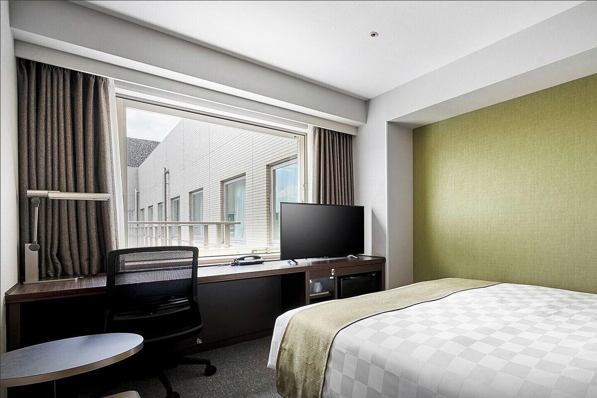 Quarto de hotel com cama, escrivaninha, cadeira, televisão e janela com cortinas.