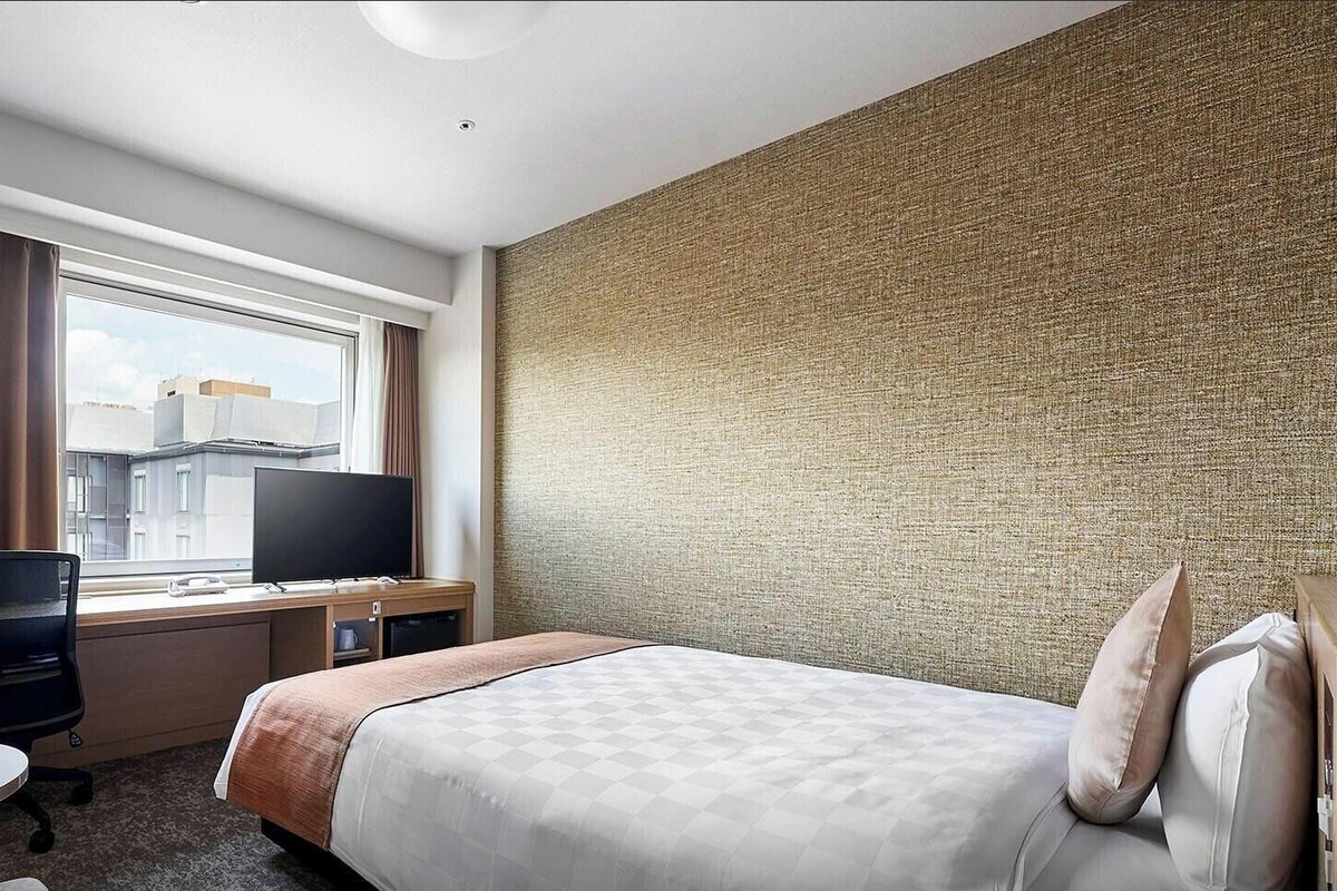 Quarto de hotel com cama, escrivaninha, cadeira e janela com cortinas.
