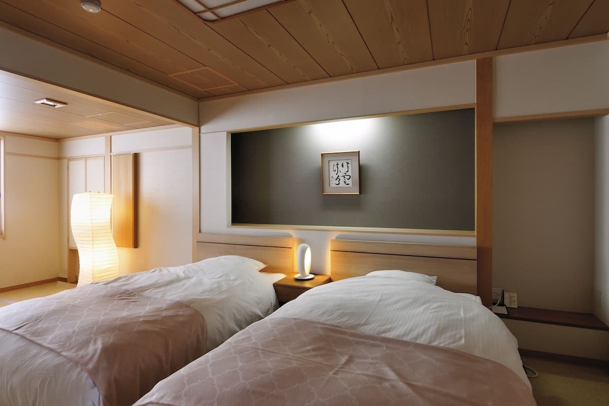 Quarto de hotel com duas camas, uma mesinha de cabeceira com abajur, televisão na parede e um quadro emoldurado.
