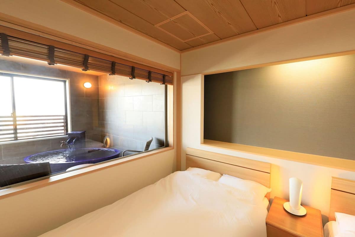Quarto de hotel com cama, mesa de cabeceira, luminária, janela com persianas e vista para uma banheira de hidromassagem.