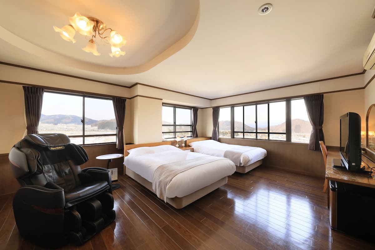 Quarto de hotel com cadeira de massagem, duas camas, uma mesa de trabalho e janelas amplas com vista para as montanhas.