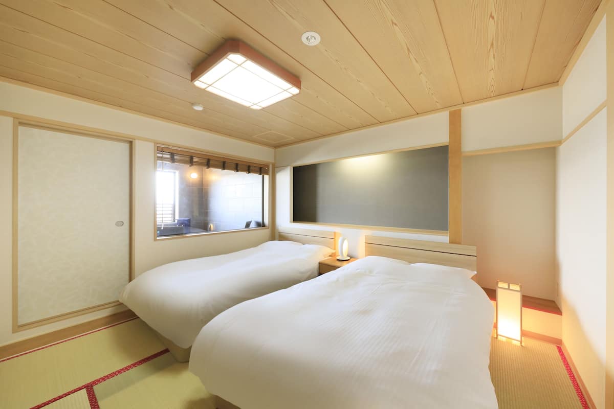 Quarto de hotel com duas camas, uma janela com cortinas, mesa de cabeceira com abajur e luminária no teto.