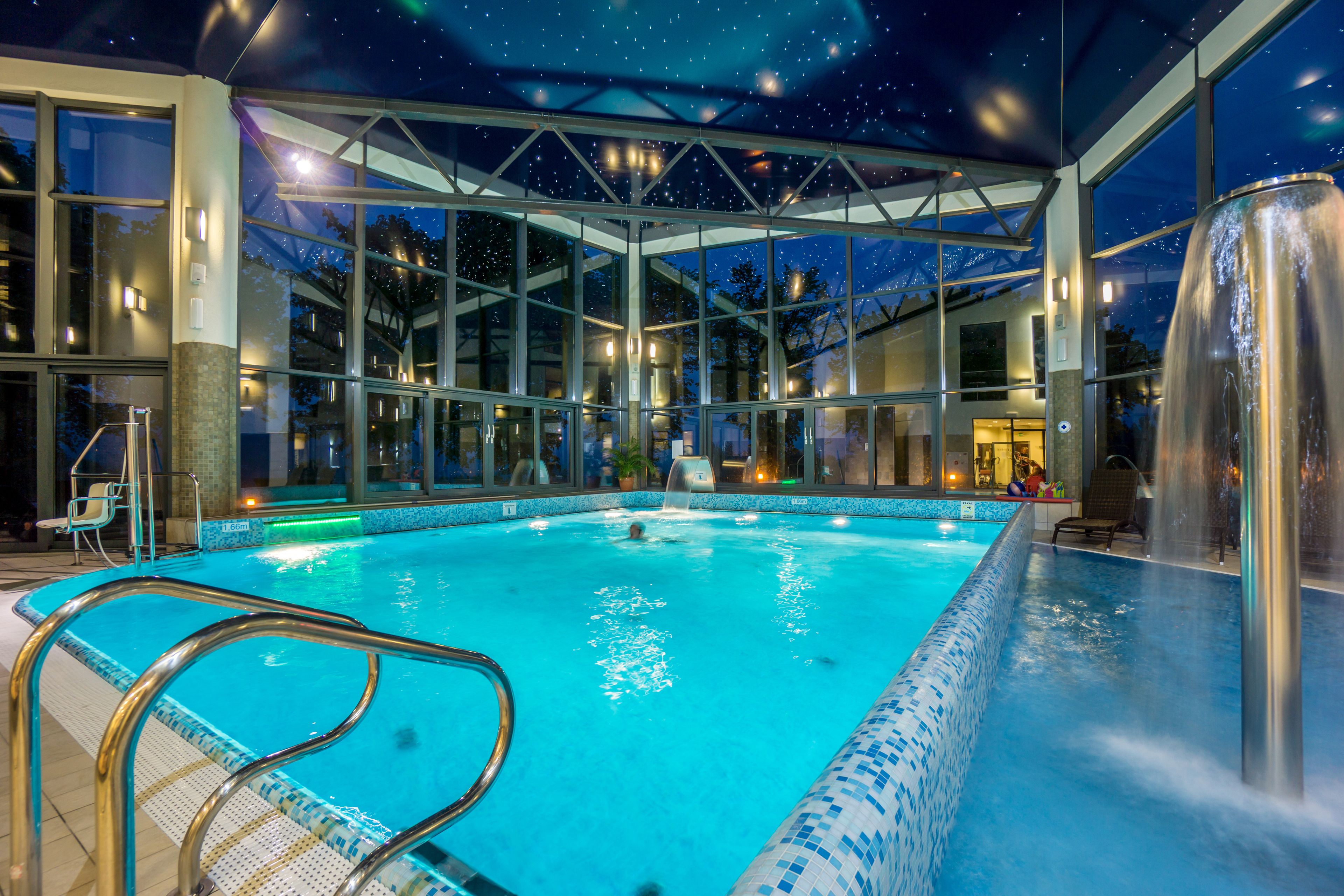 Piscina interna com teto de vidro, cadeiras confortáveis à beira da piscina e um espelho d'água.