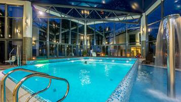 Piscina interna com teto de vidro, cadeiras confortáveis à beira da piscina e um espelho d'água.