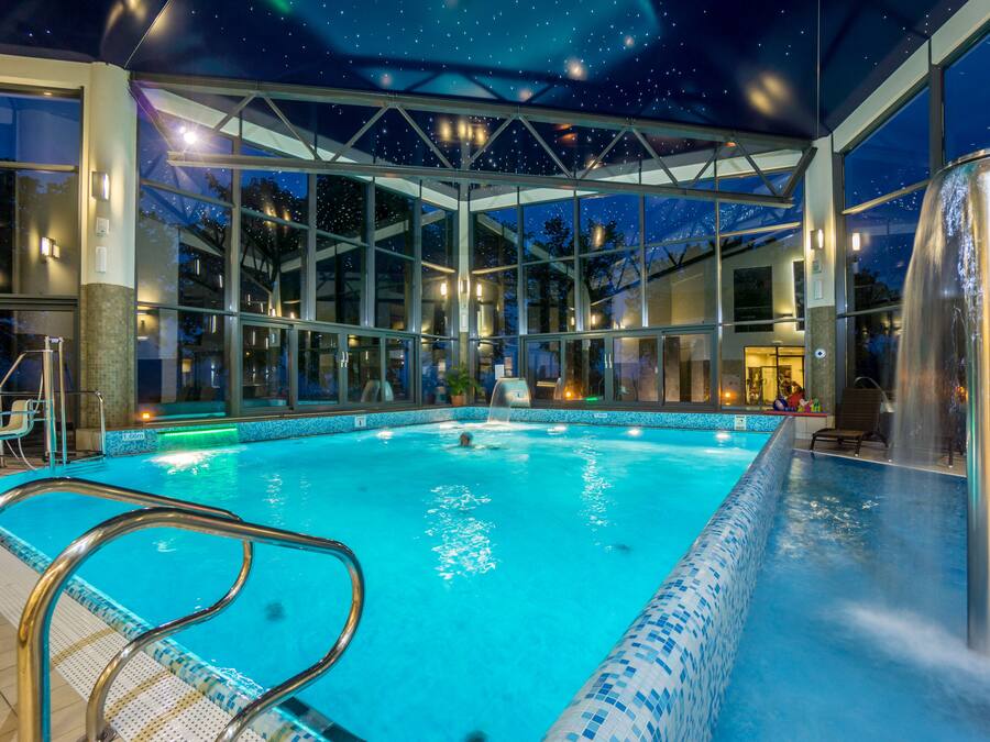 Piscina interna com teto de vidro, cadeiras confortáveis à beira da piscina e um espelho d'água.