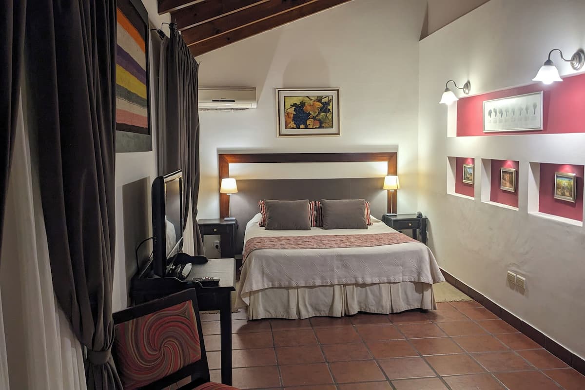 Quarto de hotel com cama, mesas de cabeceira, escrivaninha com cadeira, televisão e parede com quadros.