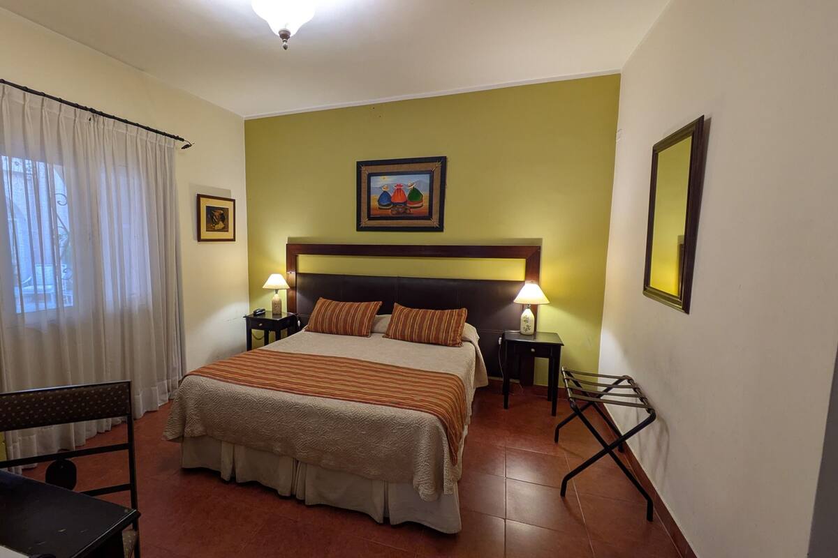 Quarto de hotel com cama, duas luminárias de cabeceira, uma cadeira, escrivaninha e janela com cortinas.