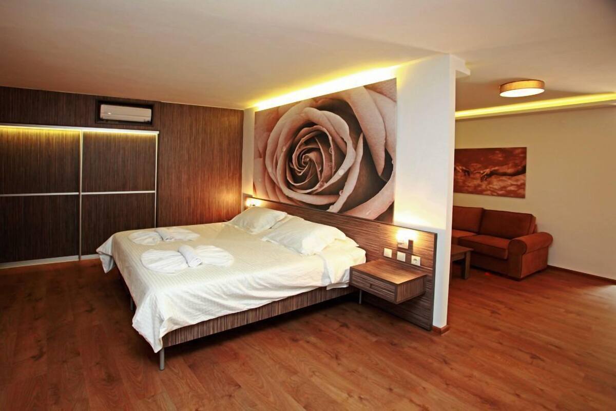 Quarto moderno com cama grande, piso de madeira, ar condicionado fixado na parede, mesa de cabeceira com luminária e uma grande obra de arte floral na parede.