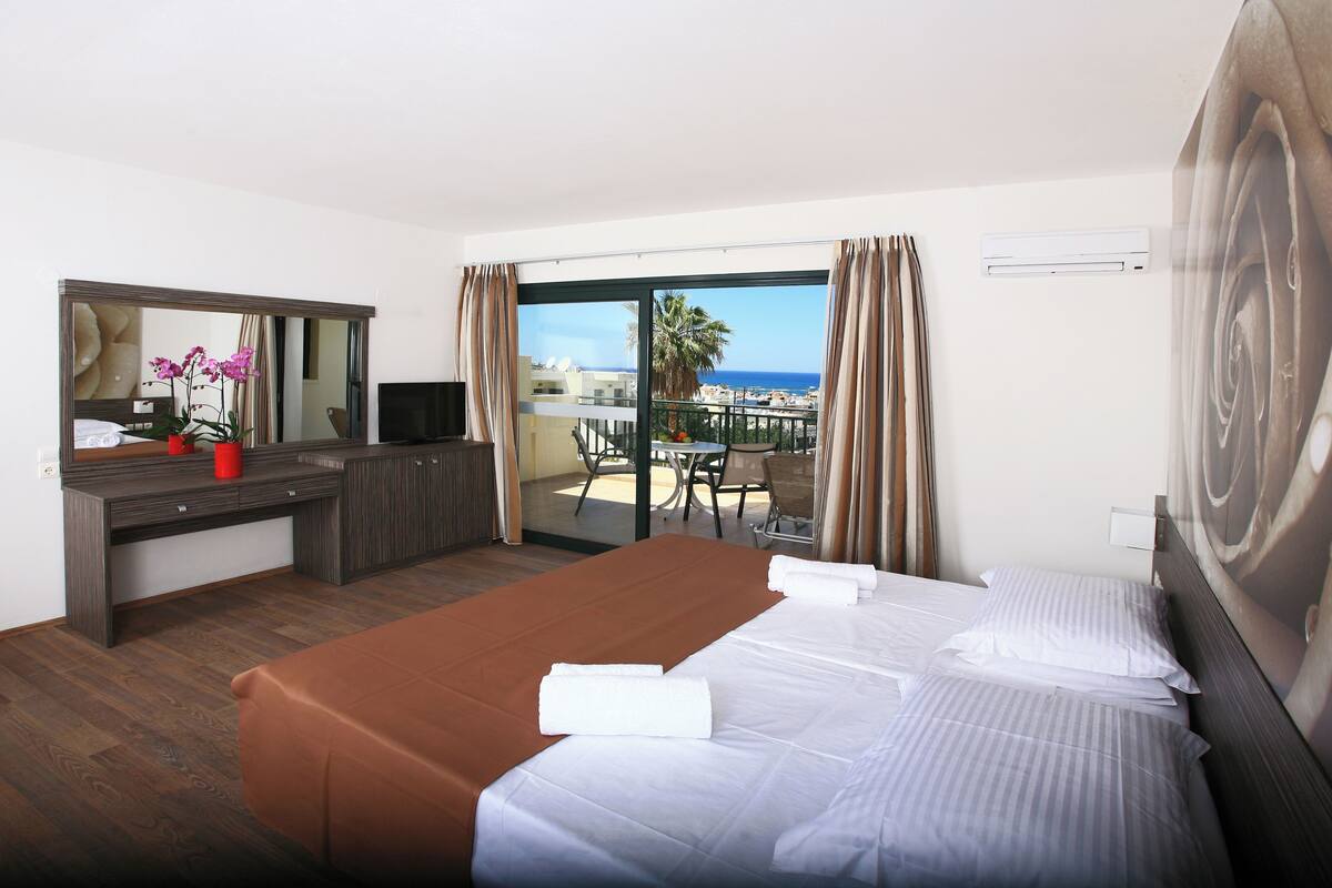 Quarto de hotel com cama, escrivaninha, televisão, varanda e vista para o mar, além de um espelho grande.
