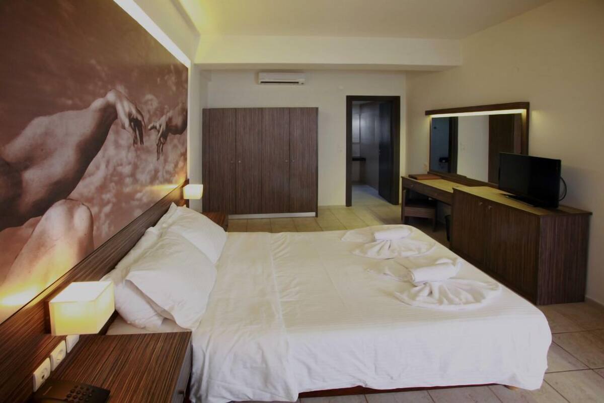 Quarto de hotel com uma cama grande, cabeceira de madeira, escrivaninha com televisão, espelho e guarda-roupa.