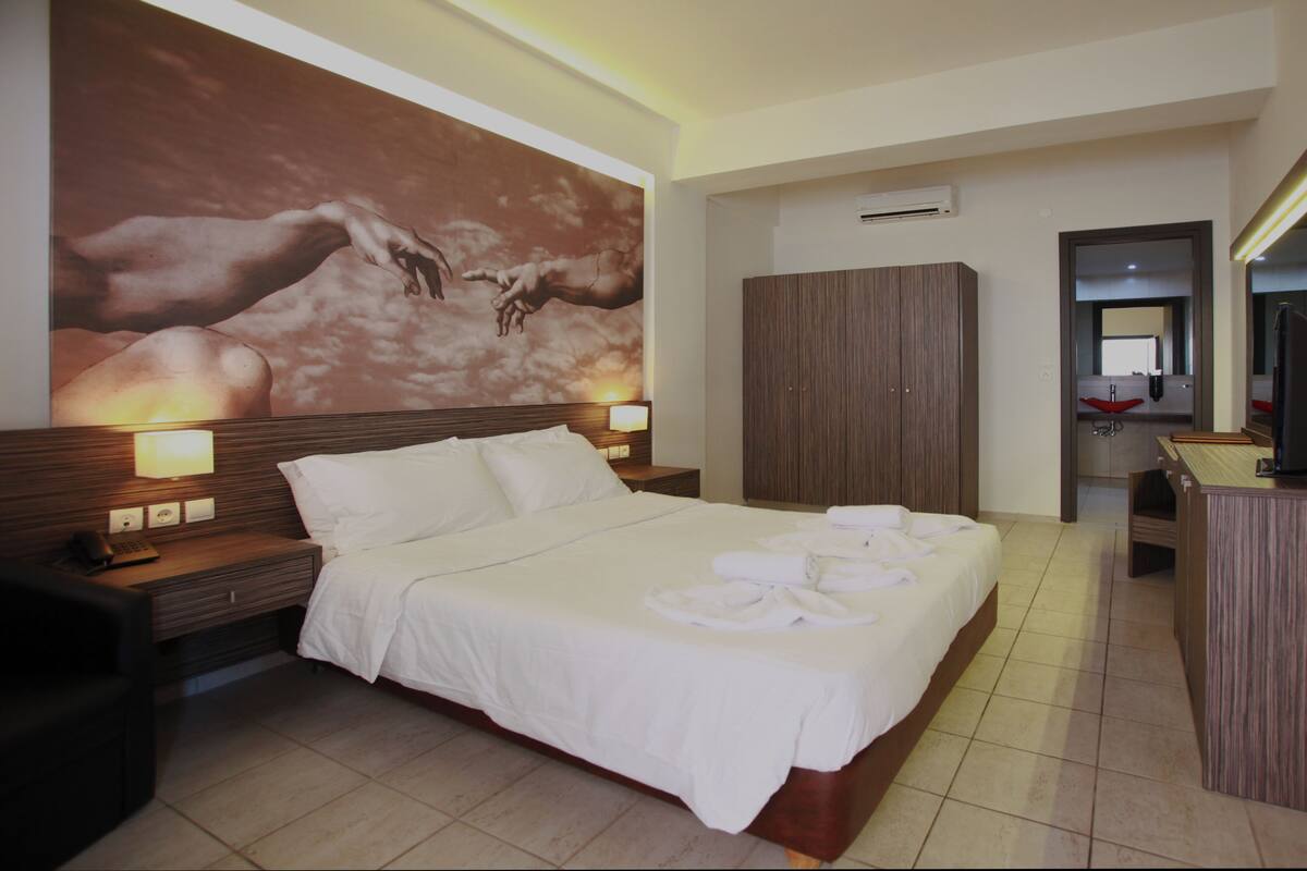 Quarto de hotel com cama grande, abajures, escrivaninha com espelho e um grande mural na parede.