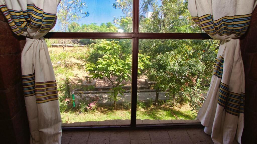 Quarto com uma janela grande, cortinas com padrões listrados e vista para um jardim com árvores e uma estrutura de pedra.